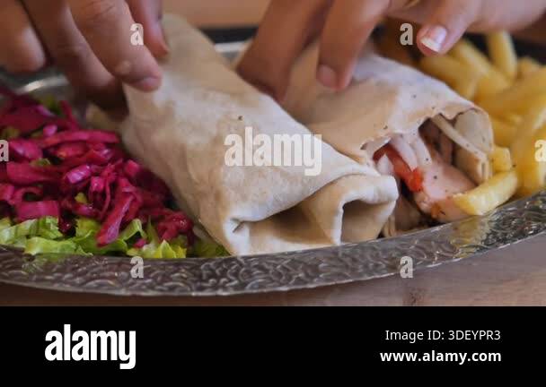 A person wraps shawarma while sitting at a table with fries and salad ...