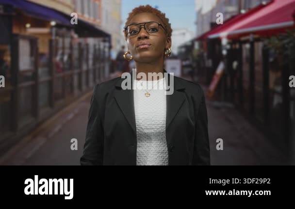 Woman with glasses and gold earrings wearing black blazer points finger to temple on street ...