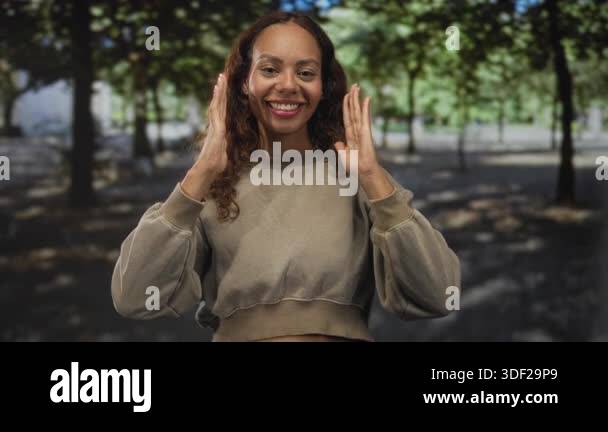 Woman framing face with hands in park among green trees smiling broadly ...