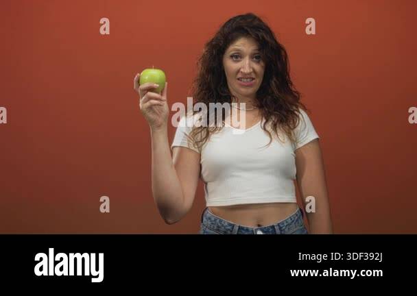 Young hispanic woman holding a green apple with raised left hand and ...