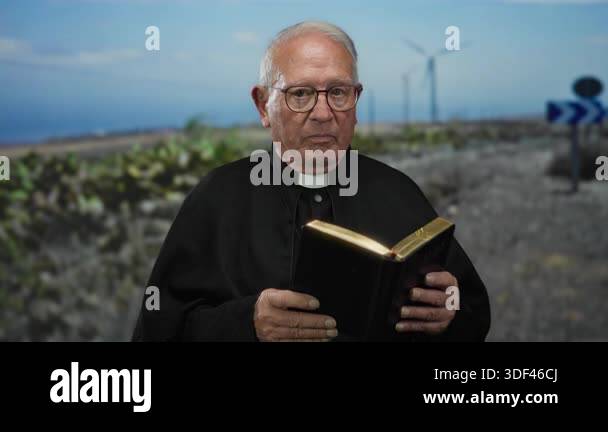 Senior priest man holding a book in an outdoor field with windmills in ...