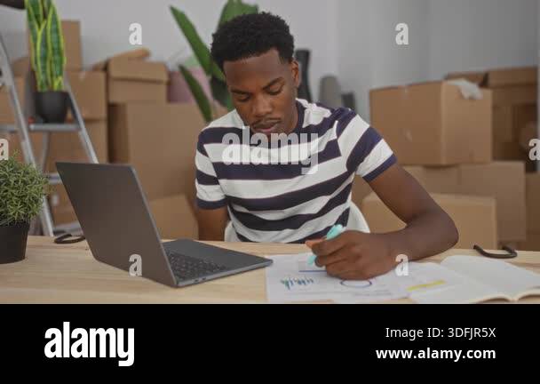 African american man seated at desk writing with pen next to laptop ...