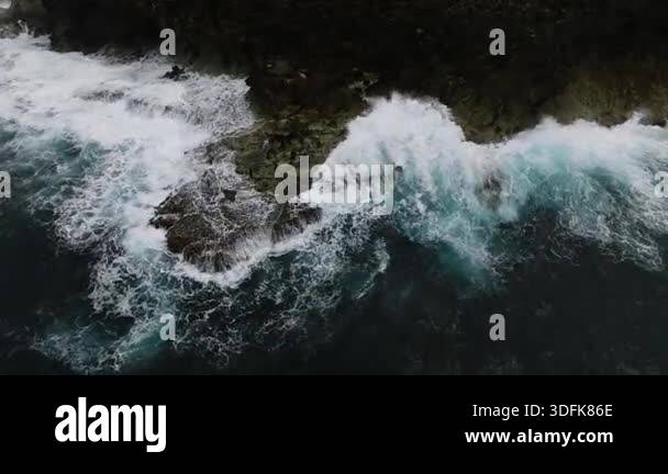 Jagged rocky cliffs are hit by strong white foamy waves under cloudy ...