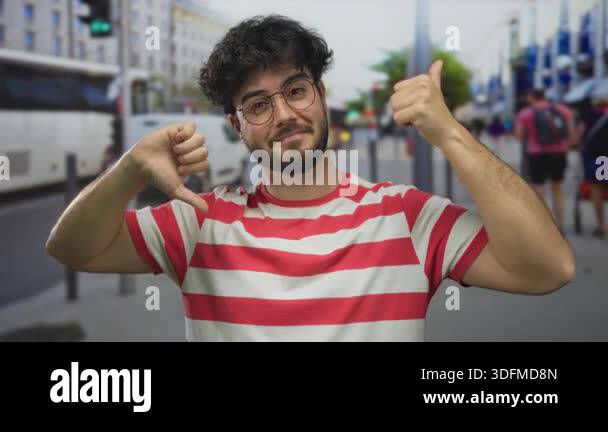 Young man with beard and glasses making thumbs up and down gestures in ...