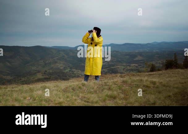 Person in yellow coat scanning valley with binoculars on grassy ridge ...