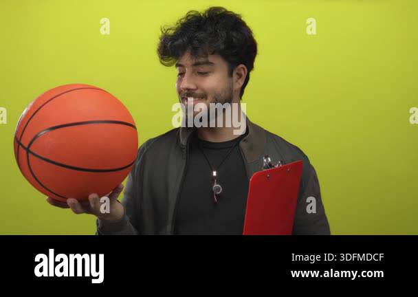 Young man with beard holding basketball and clipboard against yellow ...