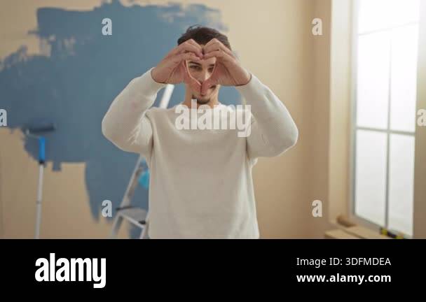 Young man making heart shape with hands indoors while painting room in ...