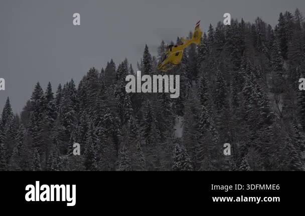 A yellow rescue helicopter flies above a snow-covered mountain forest ...