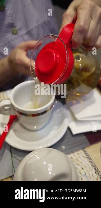 Elderly woman pouring herbal tea from a glass teapot into a white cup ...