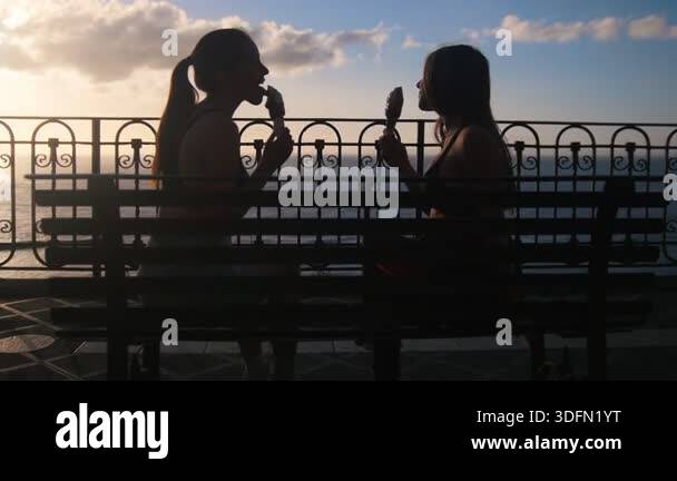 Two young women sitting on the quay of the sea and eating ice cream ...
