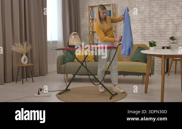 A woman using a steam iron to press a blue shirt at home, standing ...