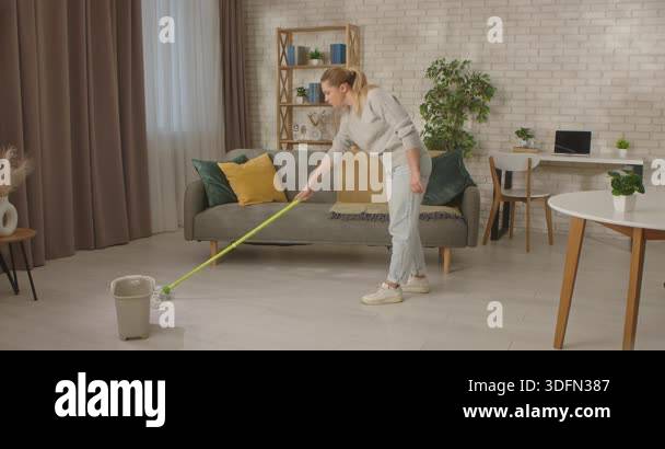 A woman is cleaning the living room by mopping the wooden floor. The ...
