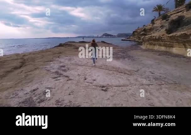 Aerial drone video of a woman running along a rocky beach by the sea ...
