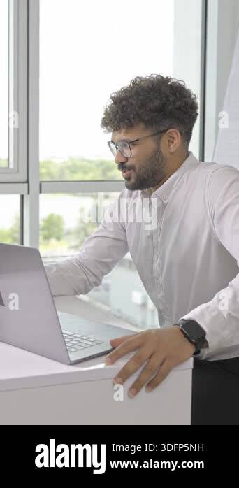 Smiling young man with glasses and curly hair working on a laptop ...