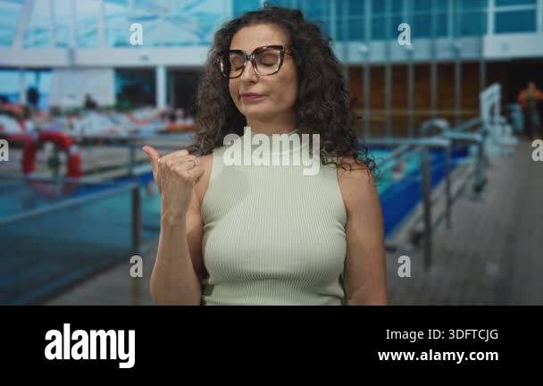 Young hispanic woman with curly hair and glasses pointing her thumb to ...