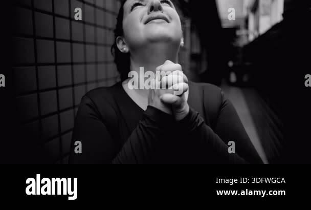 Woman clasping hands in prayer while looking upward in narrow corridor ...