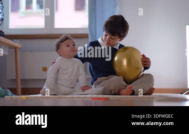 boy holding a balloon and hugging his baby brother sitting on floor at ...
