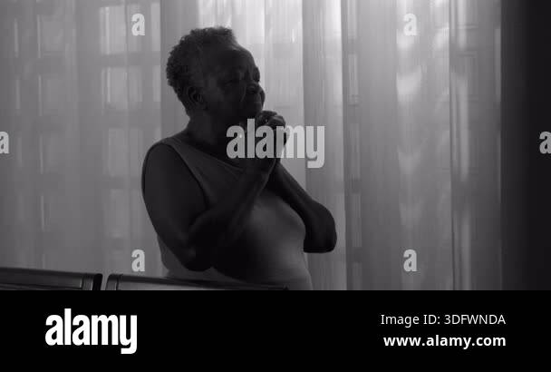 Elderly African American woman standing in prayer near window ...