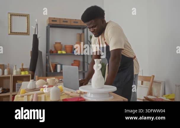 Man potter leaning over laptop at studio table with pottery wheel and ...
