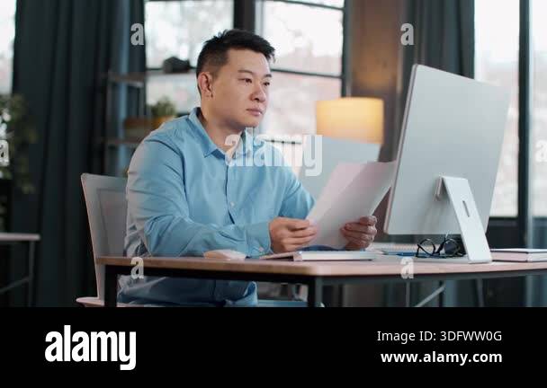 A man in a blue shirt sits at a contemporary desk, reviewing documents ...