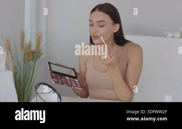 A young woman is sitting at a table, applying makeup with a brush. She ...
