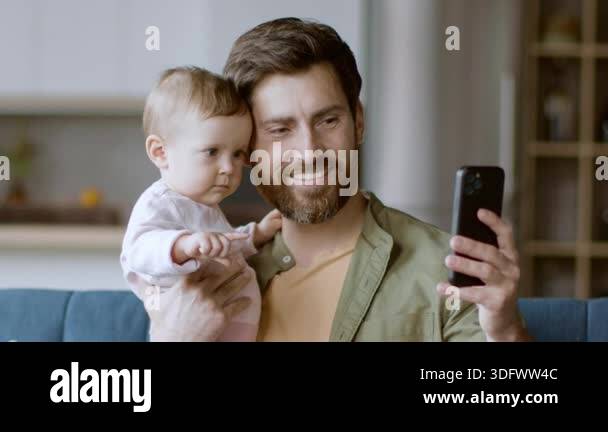 A father holds his baby while both look at a phone. They share a smile ...