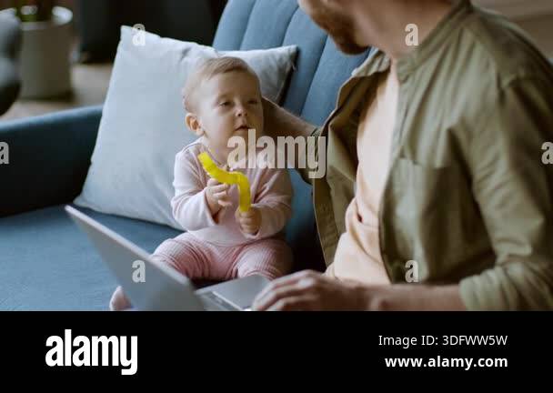 A family setting features a baby sitting on a couch, holding a banana ...