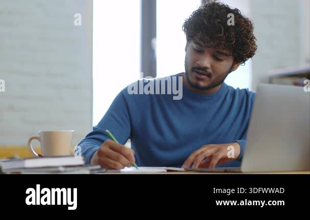 A young man with curly hair sits at a wooden desk, writing notes while ...