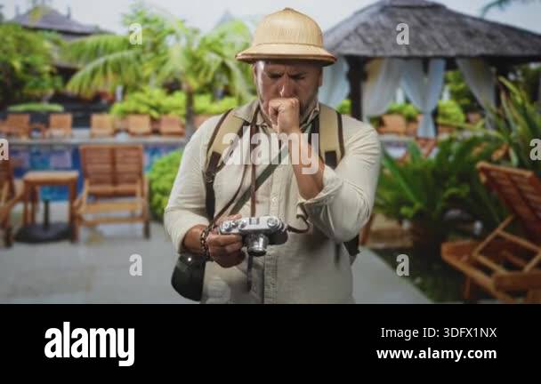 Man wearing pith helmet and backpack holding vintage camera coughs into ...