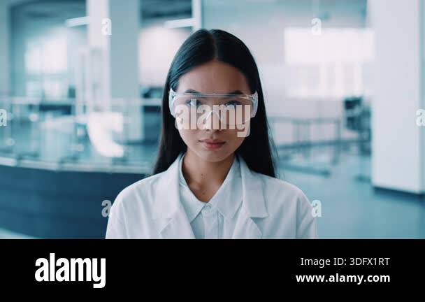 A young Asian woman in a lab coat and safety goggles focuses on her ...