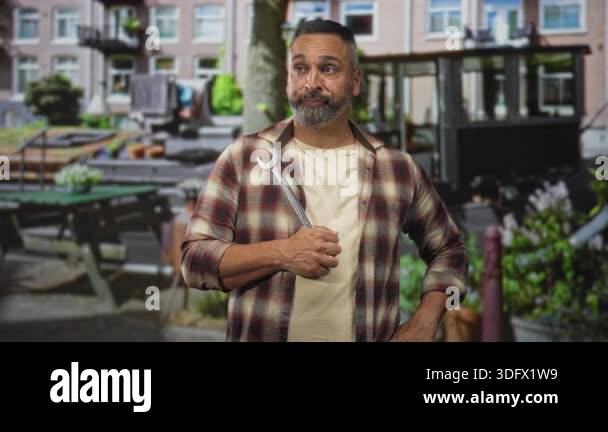 Man with grey hair and beard holds wrench to chest on a street by a ...