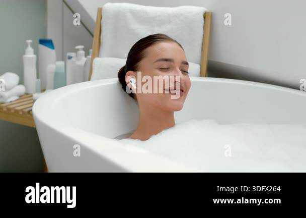 A woman is peacefully soaking in a bubble bath, wearing headphones. She ...