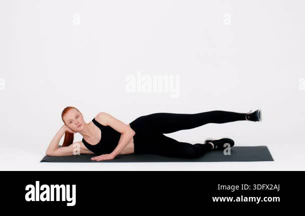 A young woman with red hair is exercising on a mat, performing side leg ...