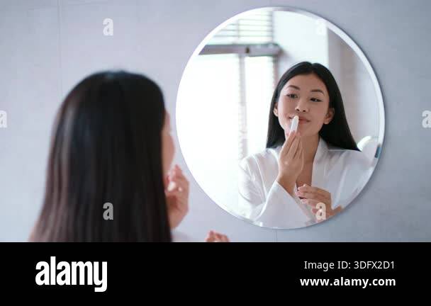 A young Asian woman applies lip care products in her bathroom while ...