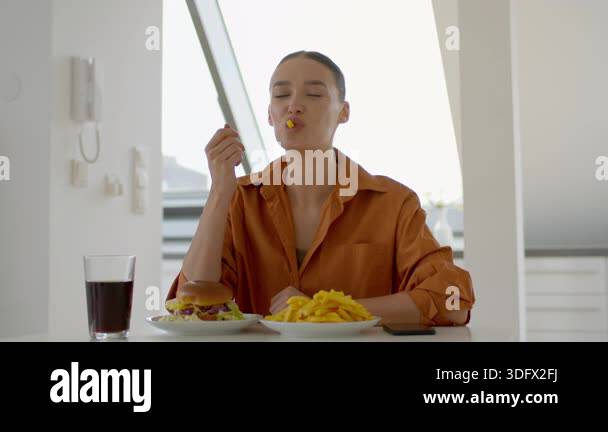 A woman sits at a table in her home, enjoying a burger and fries. She ...