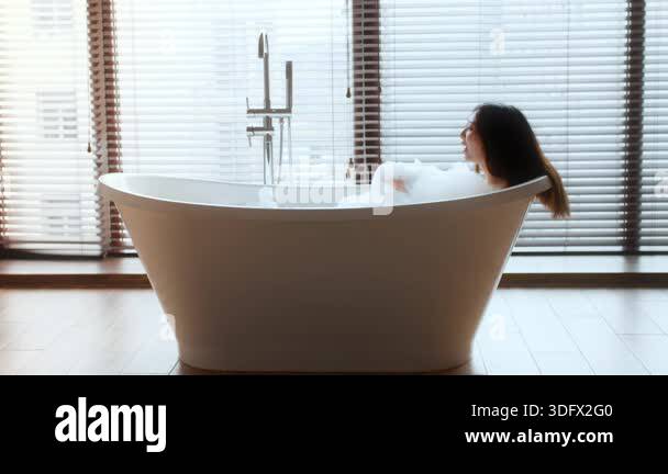 A young Asian woman relaxes in a bubble-filled bathtub, embracing self ...