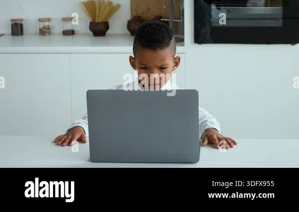 A boy sits alone at a table, focused on a laptop. He wears a white ...