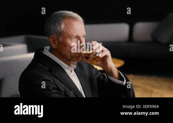 An elderly man in a suit sips a drink while reflecting in a ...