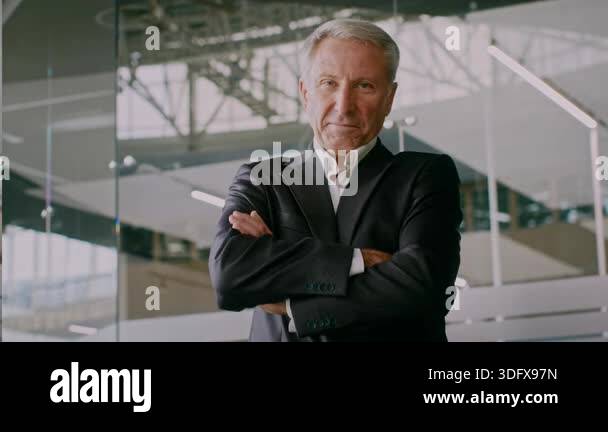 An accomplished senior man in a business suit stands with arms crossed ...