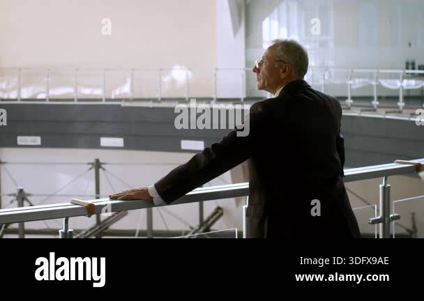 An elderly man in formal attire stands thoughtfully at a glass railing ...