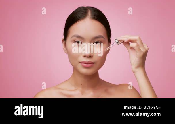 A young Asian woman applies a skincare serum to her face, showcasing ...