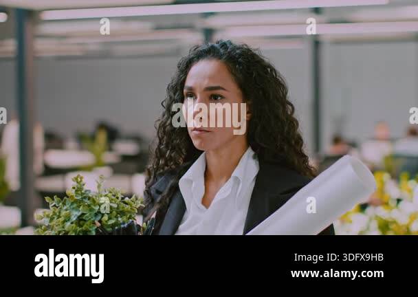 A manager with curly hair carries a small plant and rolled documents in ...