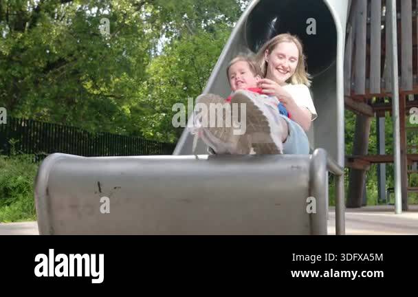 A happy mother and her toddler son slide down a slide at a modern city ...