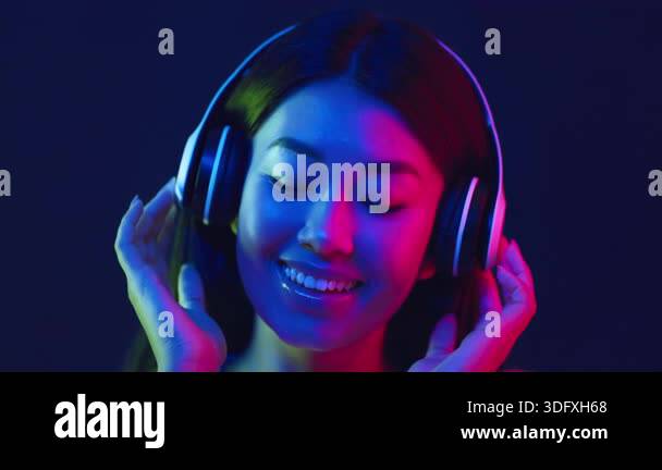 A young Asian woman listens to music with headphones, smiling in a ...