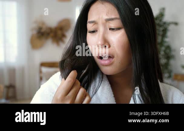 A young woman of Asian descent is at home, focusing on her hair while ...