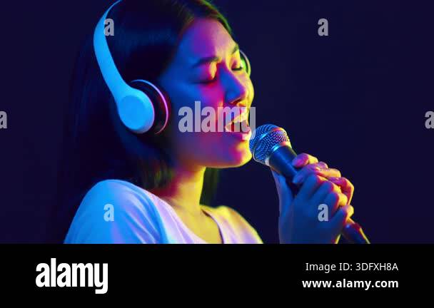 A young Asian woman sings into a microphone while wearing headphones ...
