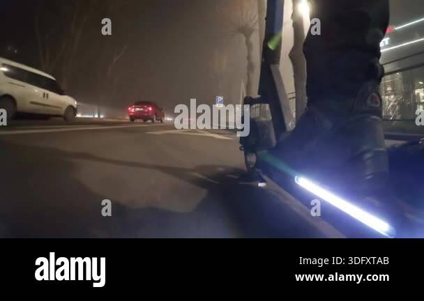A scooter rides along a foggy city road at night, viewed from the ...