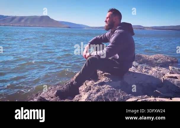 Young attractive traveler sitting on the rocky shore of a mountain lake ...