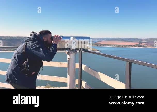 Young attractive guy looking through binoculars at a mountain lake from ...