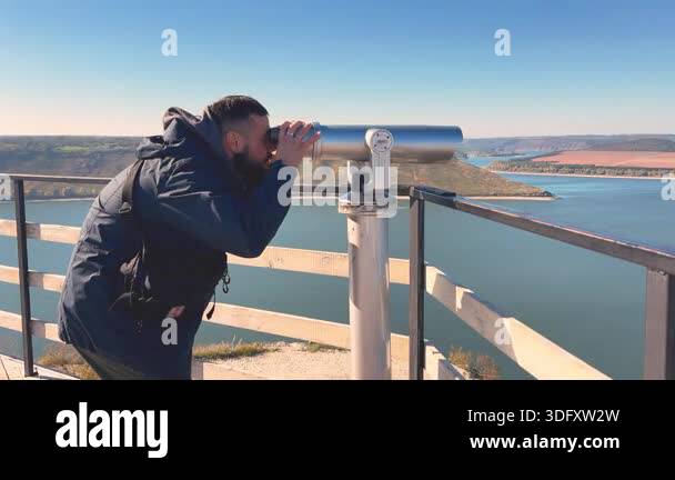 Young attractive guy looking through binoculars at a mountain lake from ...